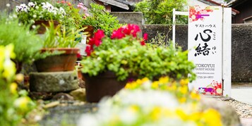 Um jardim com diversas plantas e flores em vasos, uma placa branca com caracteres japoneses e um muro de pedra.