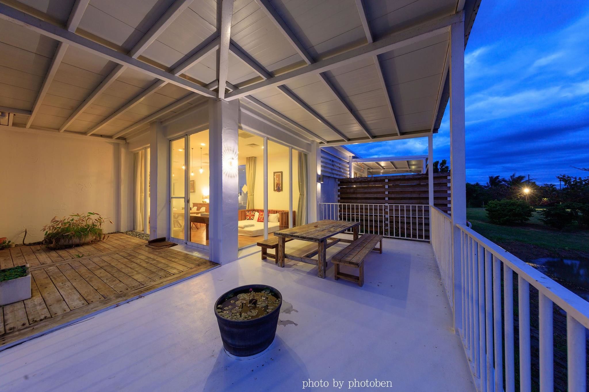 Uma área externa coberta com deck de madeira, uma mesa e bancos, e um vaso de planta.