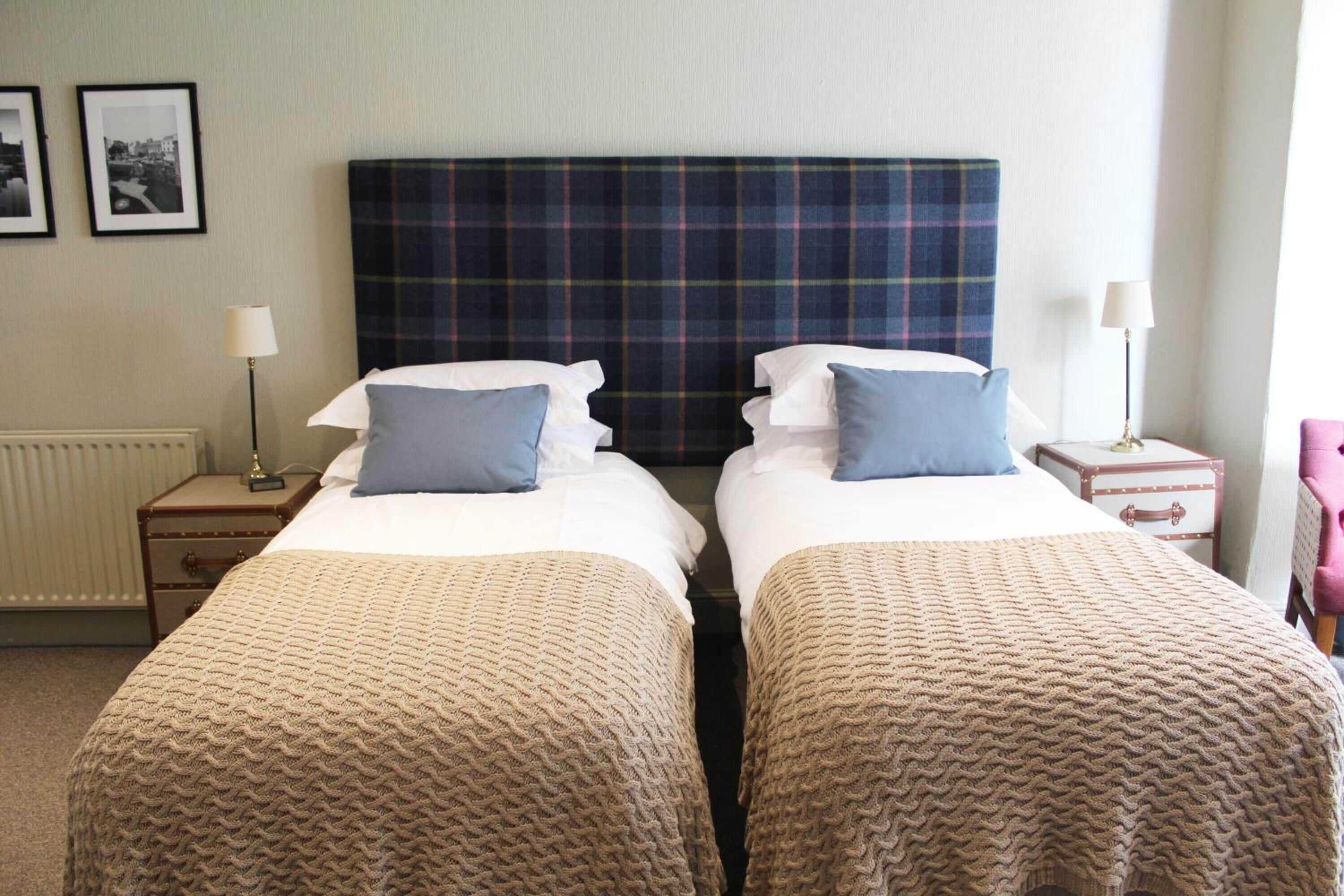 Quarto de hotel com duas camas, cabeceira xadrez e uma mesinha de cabeceira com abajur.