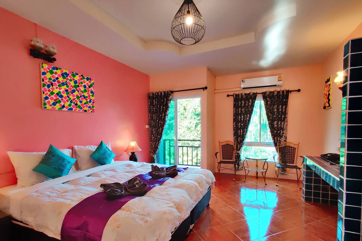 Quarto de hotel com cama, uma pequena mesa, duas cadeiras, janela com cortinas, ar condicionado e uma obra de arte colorida na parede.