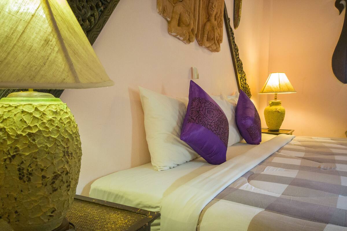 Uma cama bem arrumada com colcha xadrez, um travesseiro com padrão floral roxo e uma luminária em uma mesa de cabeceira.