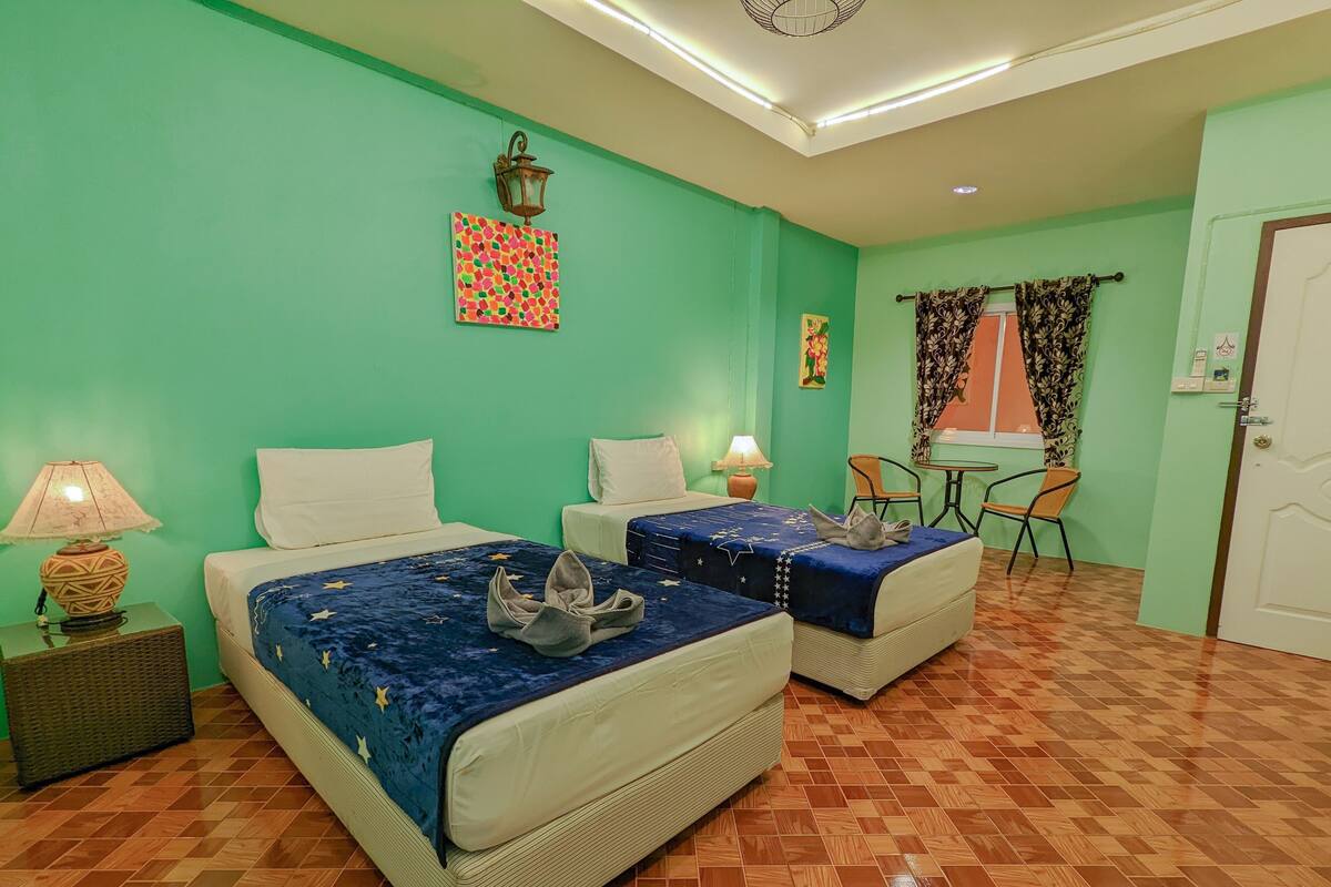 Quarto com duas camas, uma mesinha, cadeiras, um abajur e um objeto decorativo na parede.
