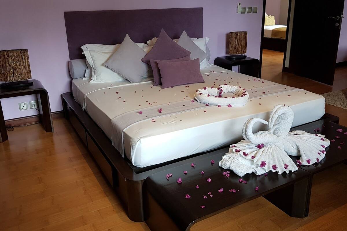 Uma cama bem arrumada com toalha e pétalas de rosa, uma mesa de cabeceira e vista para outro quarto.