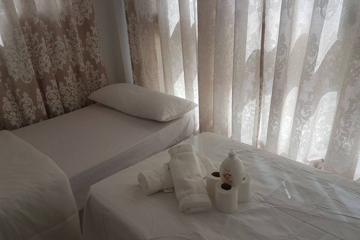 Quarto de hotel com duas camas, banheiro e janela com cortinas leves.