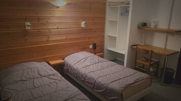 Quarto com duas camas de solteiro, cabeceira de madeira, mesa de cabeceira, prateleira com abajur e escrivaninha com cadeira.