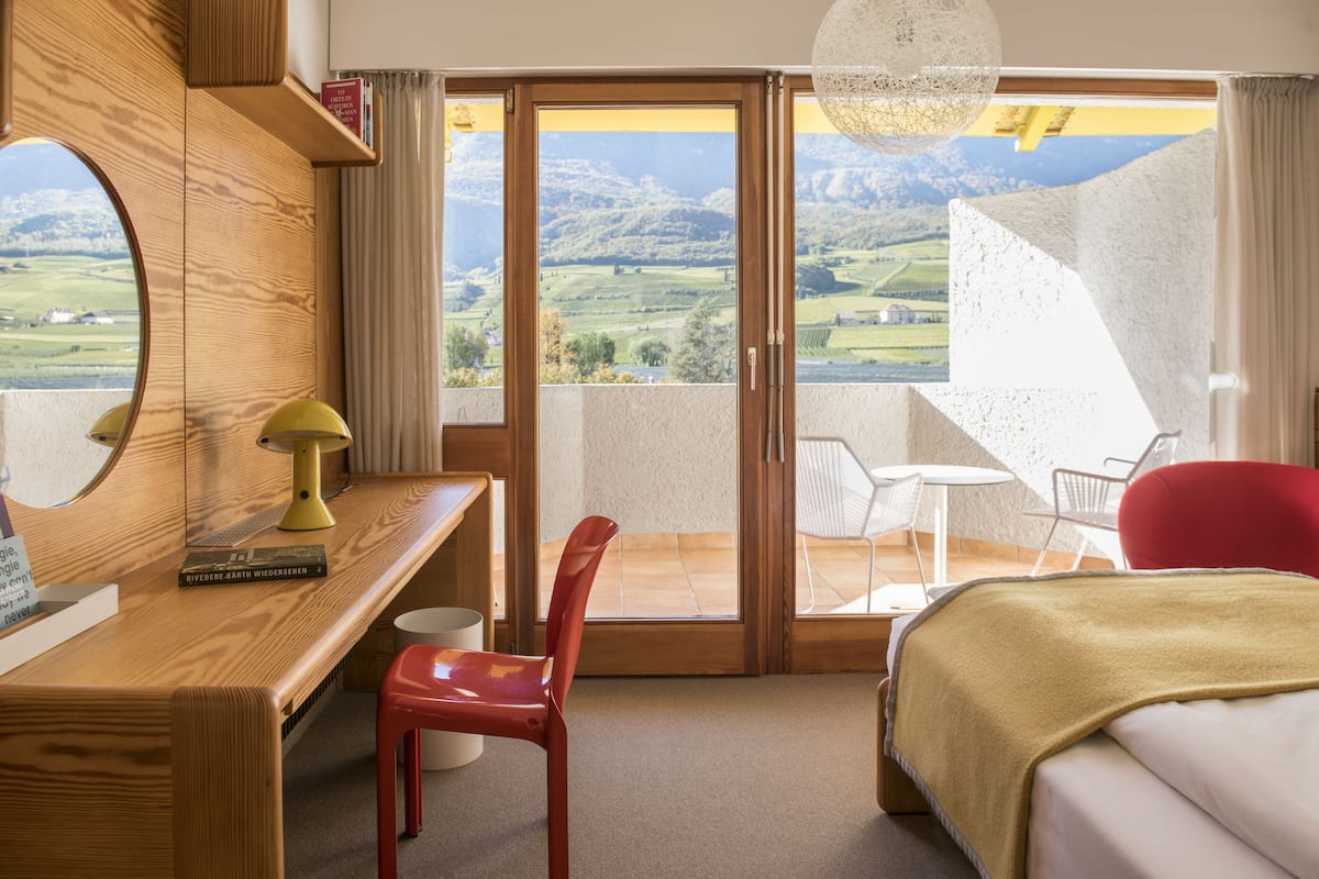 Quarto de hotel com escrivaninha de madeira, uma cadeira vermelha, cama com manta bege, uma mesa branca e vista para uma paisagem verde através de uma porta de vidro.