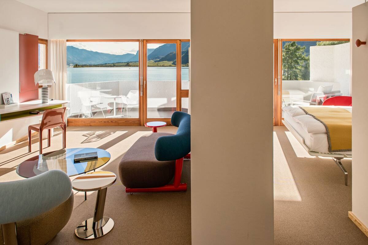 Quarto de hotel moderno com varanda e vista para um lago e montanhas.