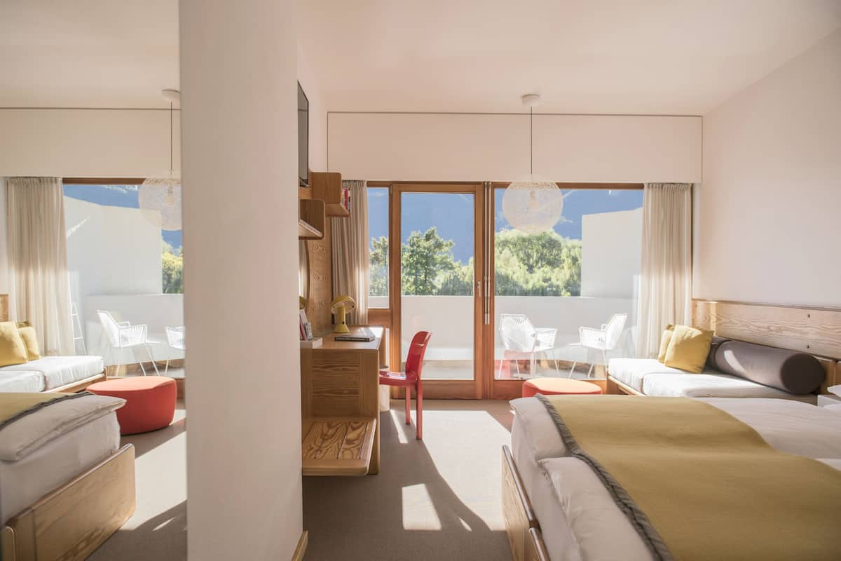 Quarto de hotel moderno com uma grande porta de vidro deslizante para a varanda, uma mesa de madeira, duas camas e um espaço de estar com cadeiras brancas.