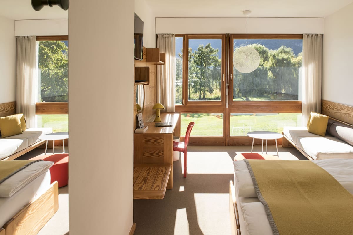 Quarto de hotel com duas camas, uma escrivaninha e uma cadeira. Janelas amplas oferecem vista para árvores e um lago.