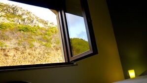 Quarto com cama, cadeira e janela com vista para terreno rochoso e vegetação.