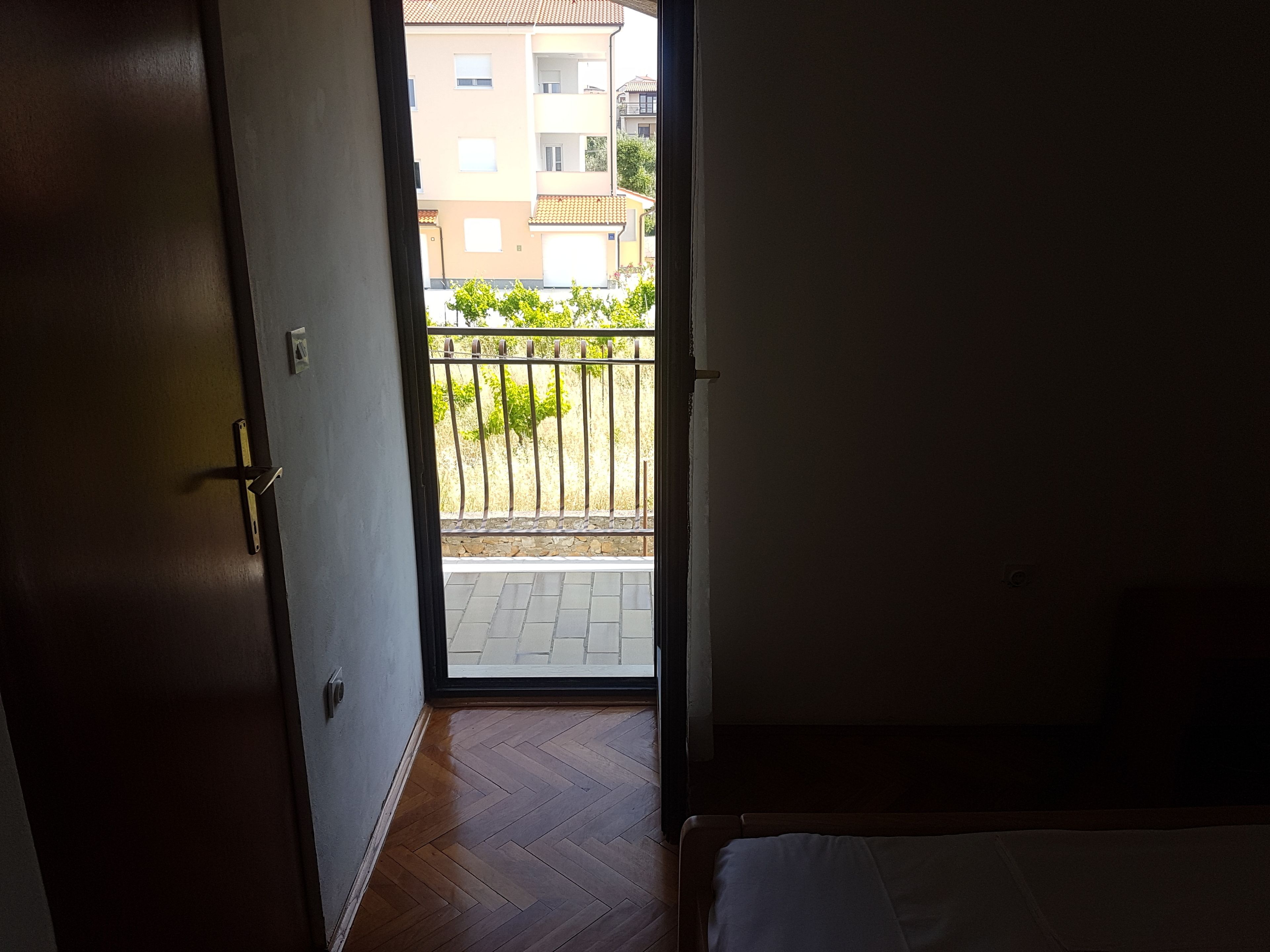 Quarto com piso de madeira, uma cama e uma porta que dá para uma varanda com vista para edifícios e uma cerca.