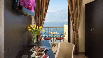 Quarto de hotel com escrivaninha, cadeira, pequena mesa com um vaso de flores amarelas, televisão na parede e vista para o mar e montanhas através das cortinas.