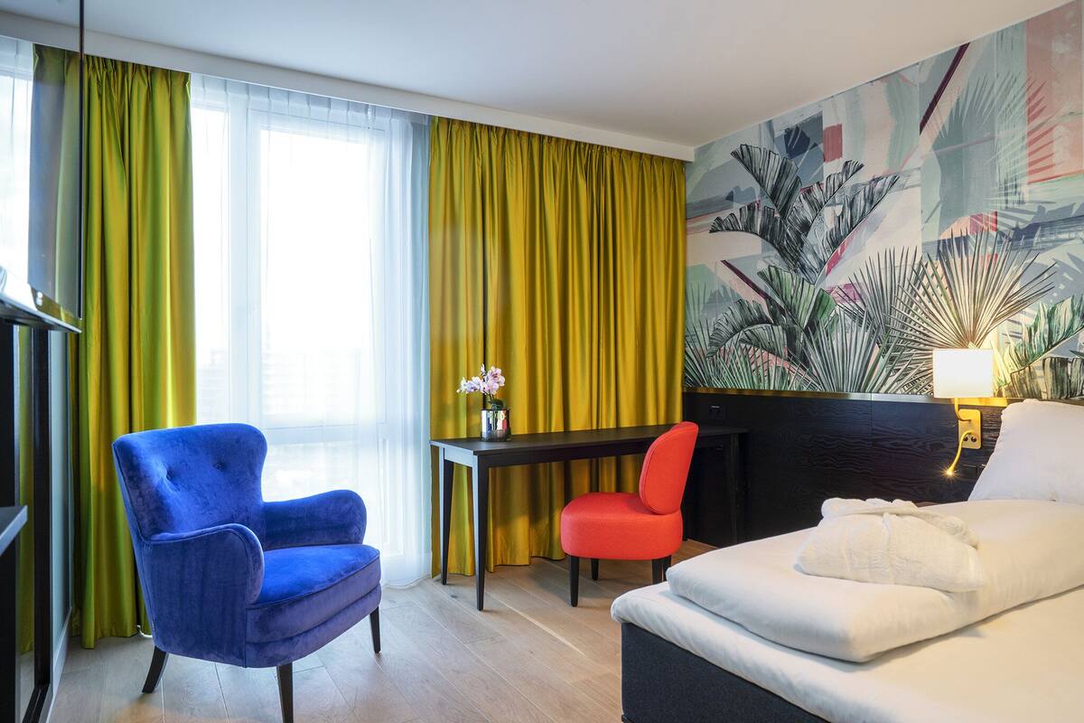 Quarto de hotel com uma cama, uma poltrona azul, uma cadeira vermelha, uma mesinha de centro com um vaso de flores e um mural na parede.