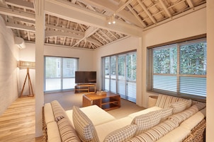 Uma sala de estar espaçosa com teto alto, janelas amplas, um conjunto de sofás, uma TV e uma mesa de centro de madeira.
