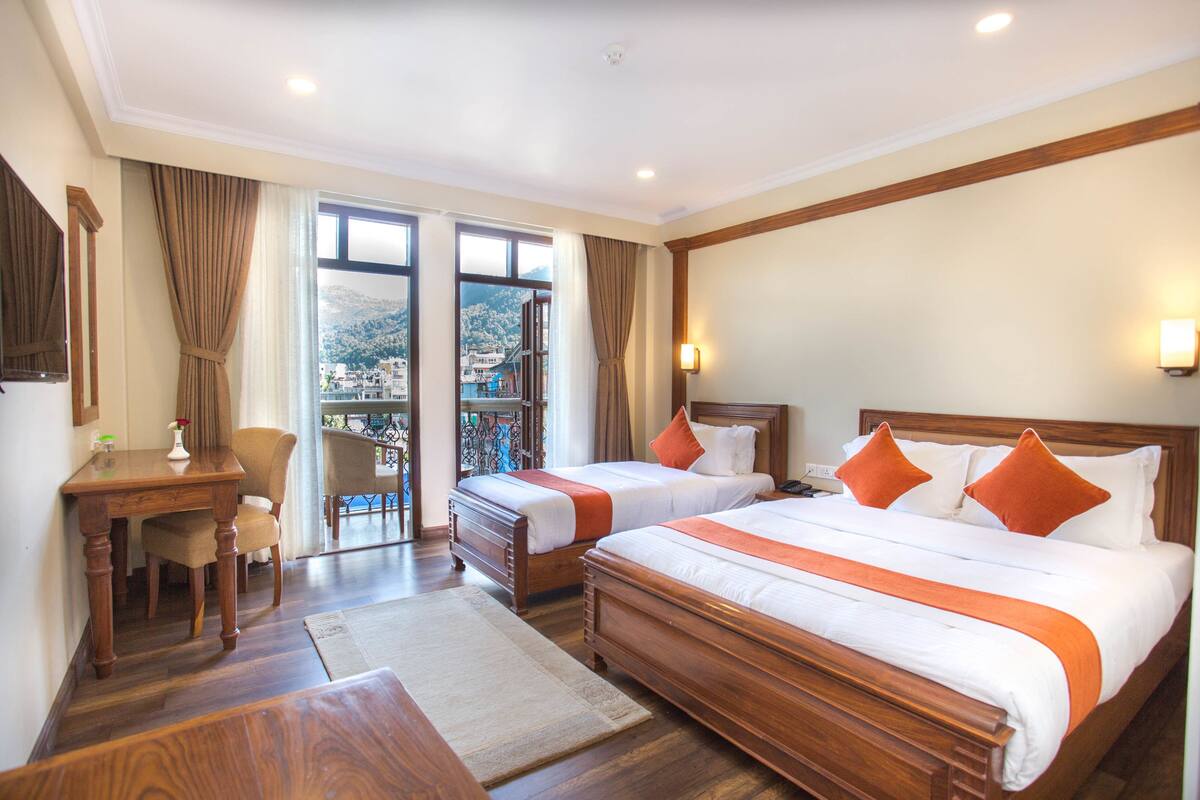 Quarto de hotel com duas camas, uma escrivaninha e uma cadeira. Janelas oferecem vista para uma paisagem montanhosa.