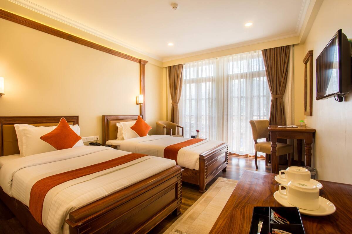 Quarto de hotel com duas camas, uma escrivaninha, uma cadeira, uma televisão e uma janela com cortinas.