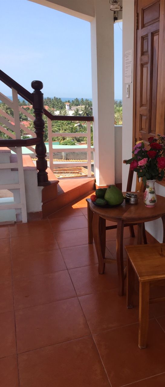 Varanda com grade de madeira e vista para o mar. Há uma mesa com um vaso de flores e um prato de frutas.