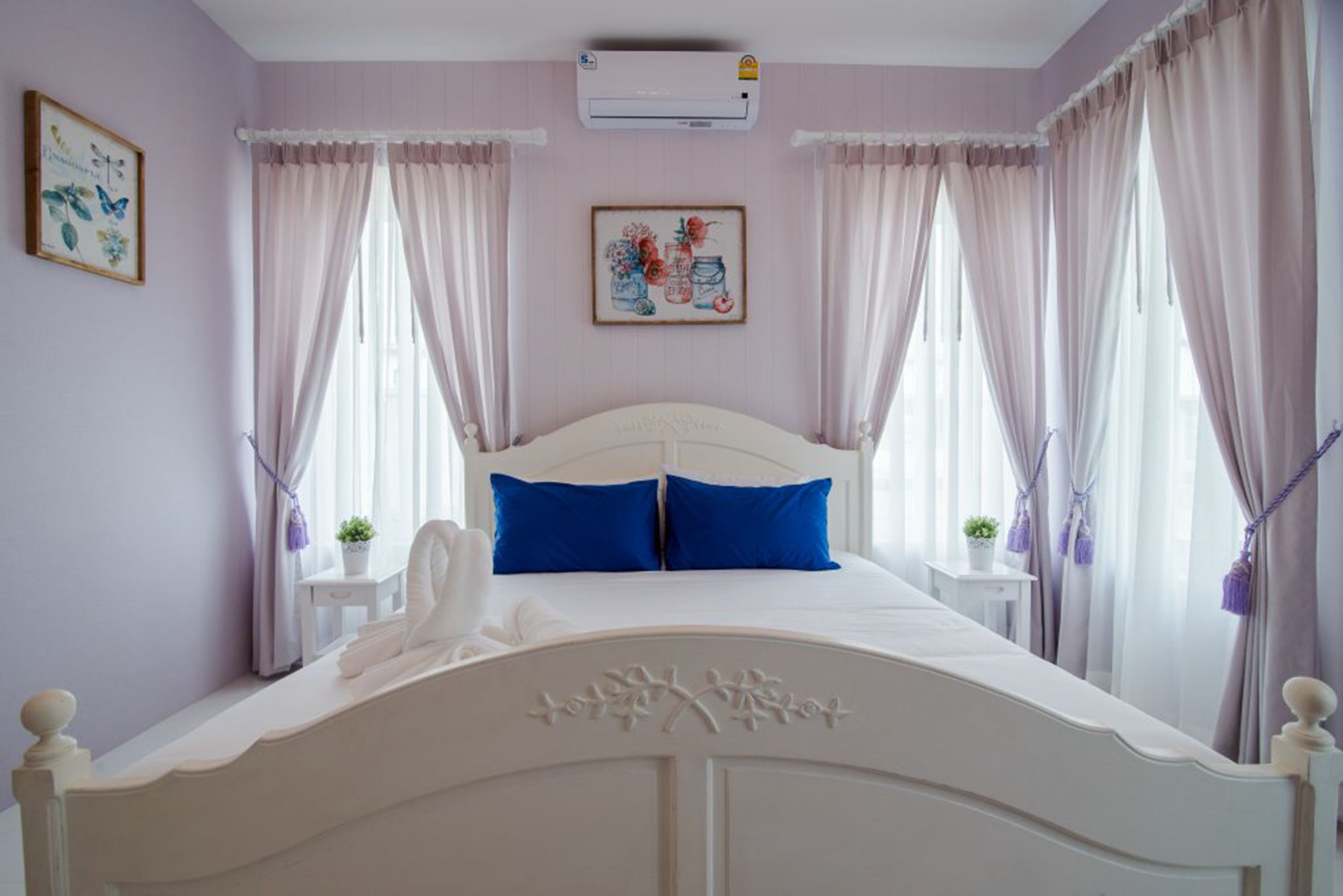 Quarto com cama branca, duas mesas de cabeceira, ar condicionado fixado na parede e duas janelas com cortinas leves.