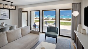 Quarto de hotel moderno com sofá bege, uma mesa redonda pequena com um vaso de flores e vista para a praia através de grandes portas deslizantes.