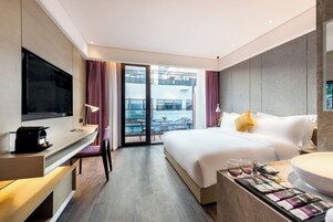Quarto de hotel com cama, escrivaninha, TV, cafeteira, uma taça de vinho e vista para o exterior.