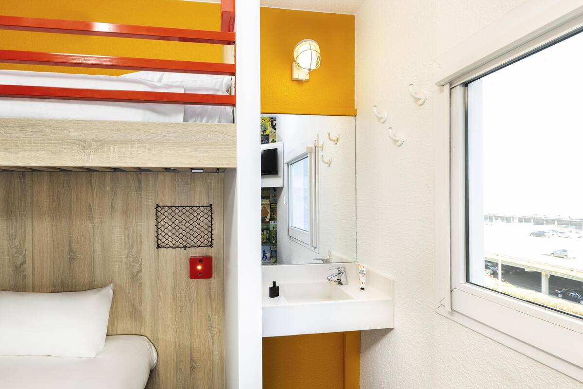 Uma cama beliche com design em painel de madeira, uma pia branca com espelho acima e uma janela com vista para um estacionamento.