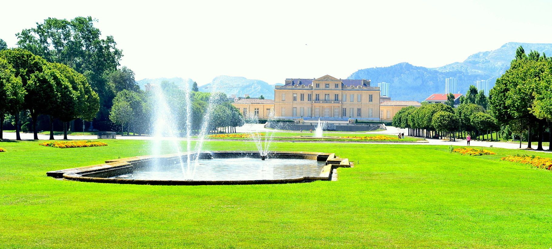 Um edifício grande com uma fonte em primeiro plano, rodeado por jardins bem cuidados e árvores.
