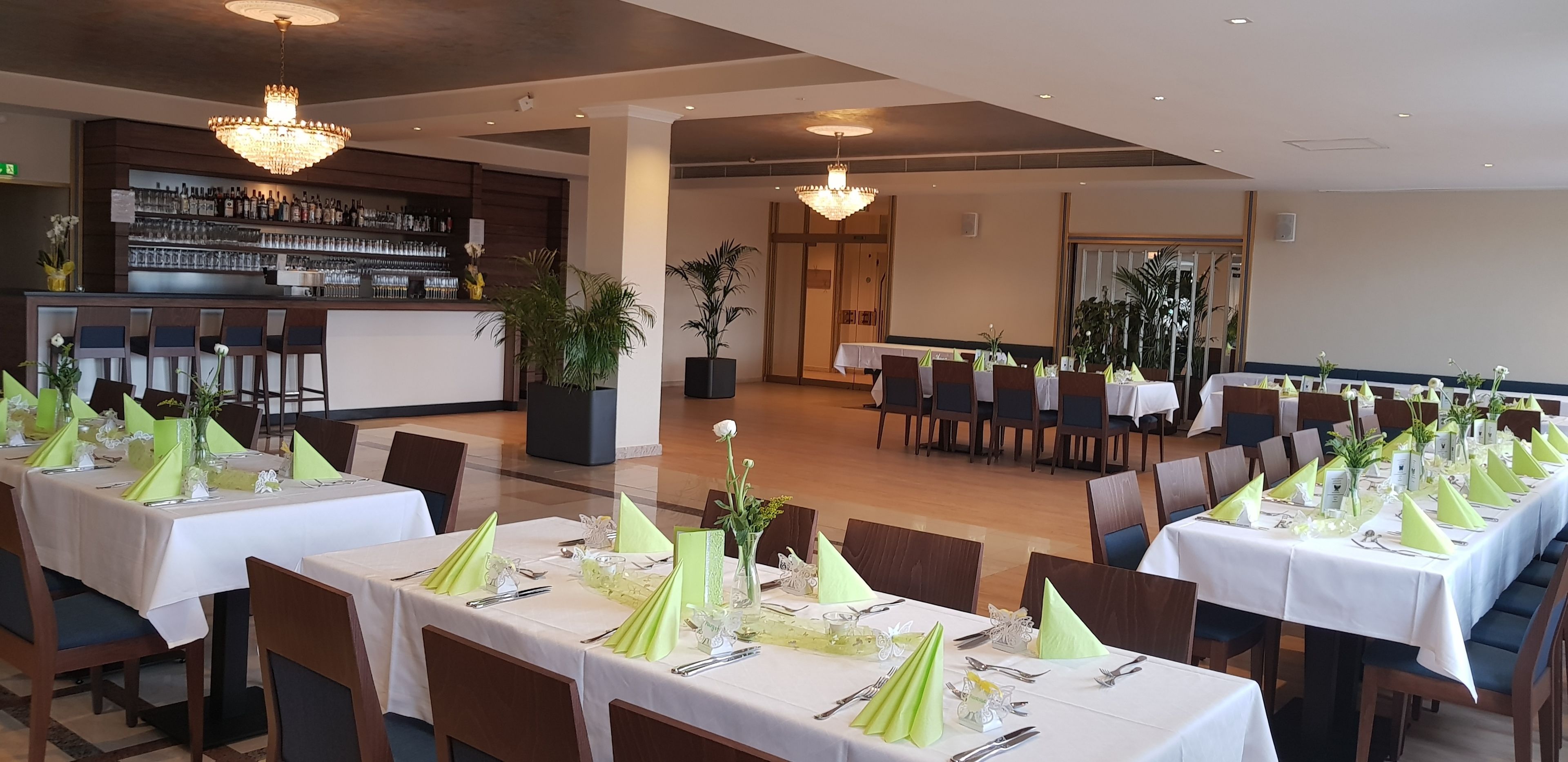 Restaurante com várias mesas preparadas para refeições, área de bar com prateleiras cheias de garrafas e grandes vasos de plantas.