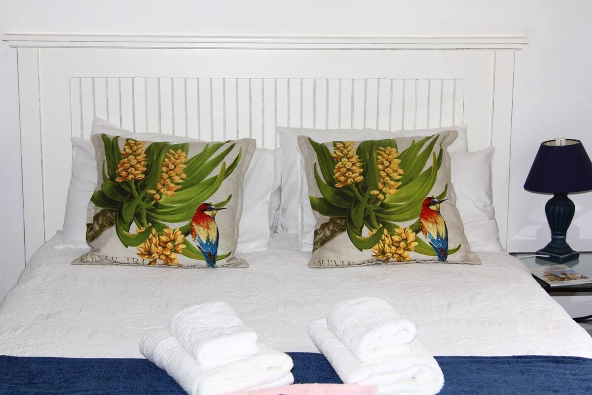 Cama com duas almofadas com padrão de pássaro e girassol, toalha branca dobrada e abajur azul escuro em uma mesa de cabeceira.