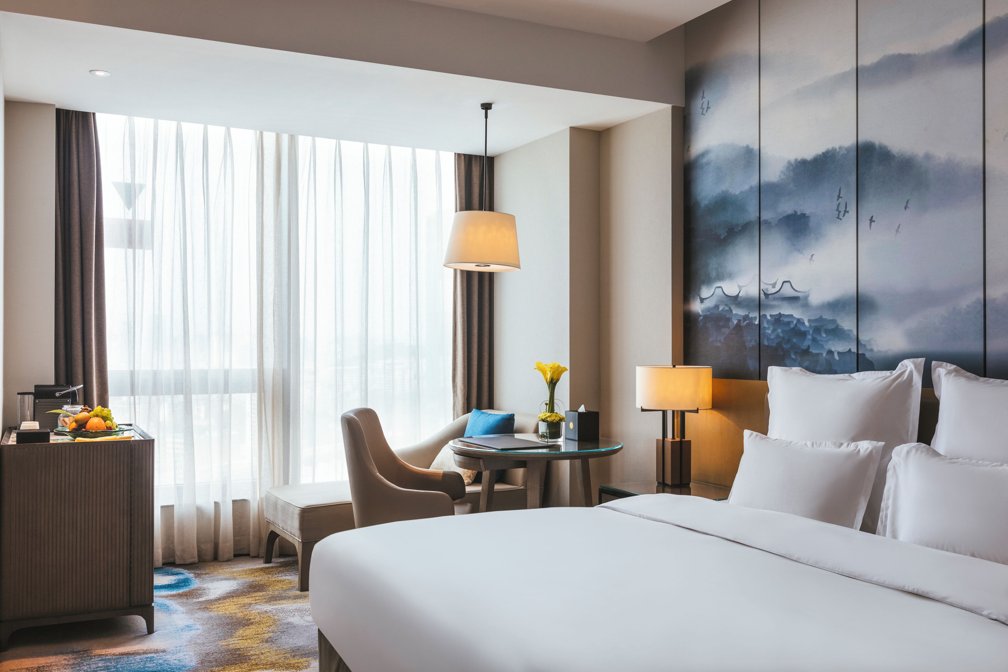 Quarto de hotel com cama, escrivaninha, cadeira, mesinha de centro com um vaso e um quadro na parede.