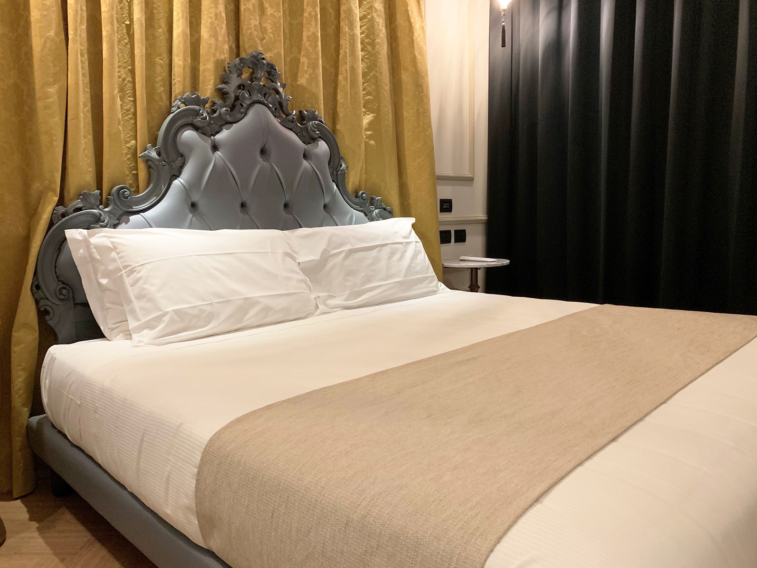 Cama bem arrumada com cabeceira estofada, roupa de cama branca e manta bege.