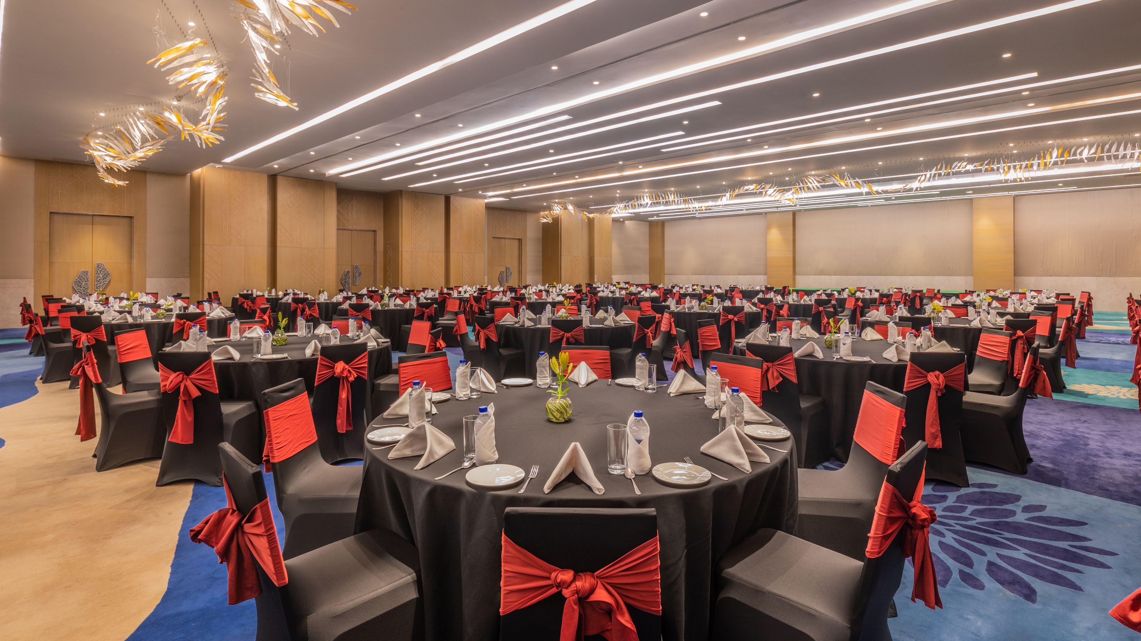 Um amplo salão de banquetes com mesas redondas, toalhas pretas e fitas vermelhas.