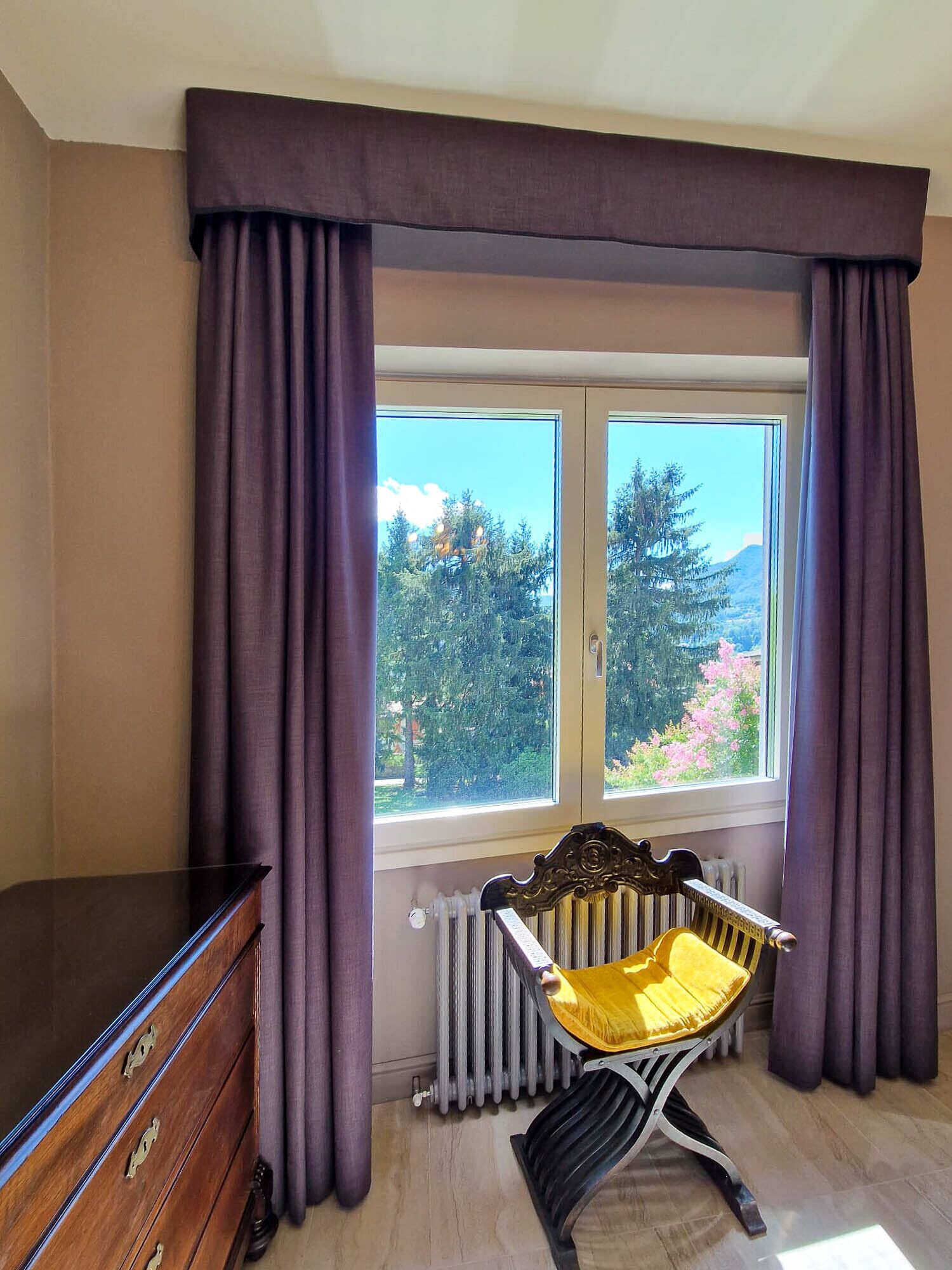 Um quarto com cômoda de madeira, poltrona amarela acolchoada e janela com cortinas roxas.