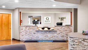 Bancada de recepção com fachada de pedra, um letreiro 'Quality by Choice Hotels' e um letreiro 'DEWAY INN', além de área de recepção com piso de madeira e tapete azul.