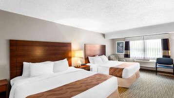 Quarto de hotel com duas camas, uma mesa de cabeceira, uma cadeira, um abajur e uma janela grande com cortinas.