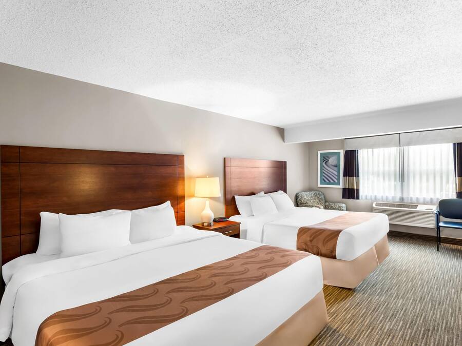 Quarto de hotel com duas camas, uma mesa de cabeceira, uma cadeira, um abajur e uma janela grande com cortinas.