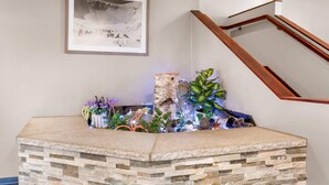 Lareira de pedra com um quadro emoldurado acima, escada de madeira à direita e um arranjo de plantas no parapeito.