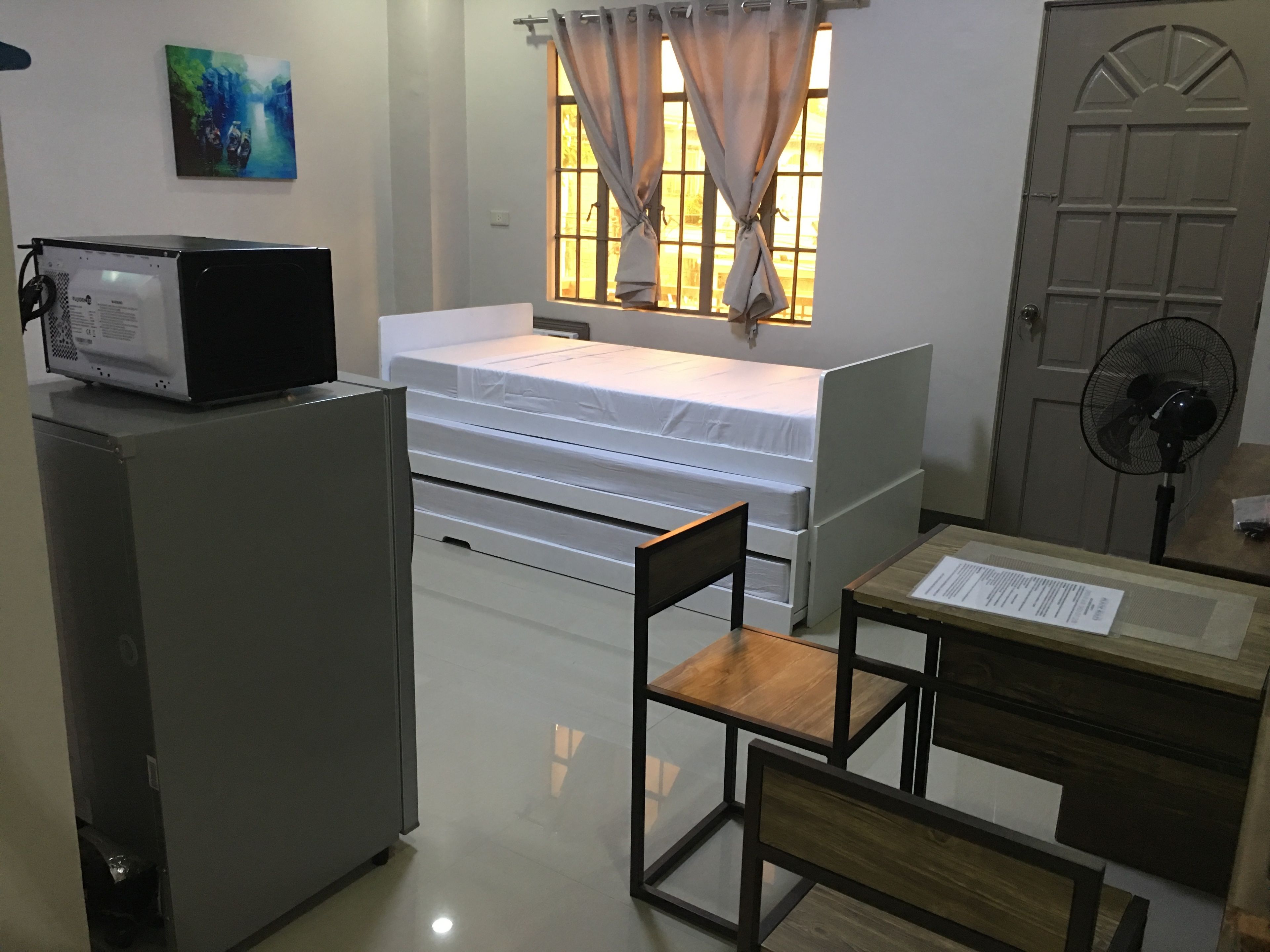 Quarto compacto com uma cama, uma mesinha com uma cadeira, um frigorífico, uma janela com cortinas e um ventilador fixado na parede.