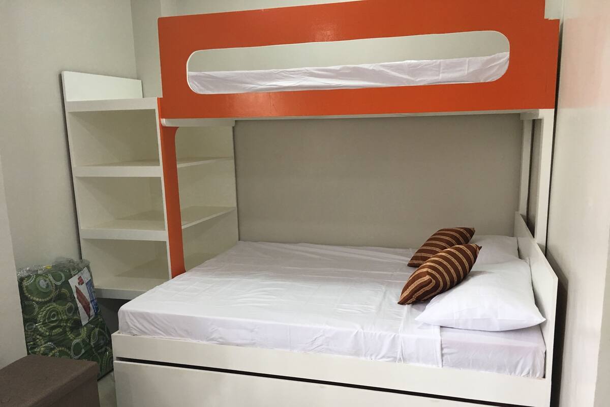 Uma cama beliche com colchão branco e almofadas listradas, cabeceira branca e uma estante à esquerda.