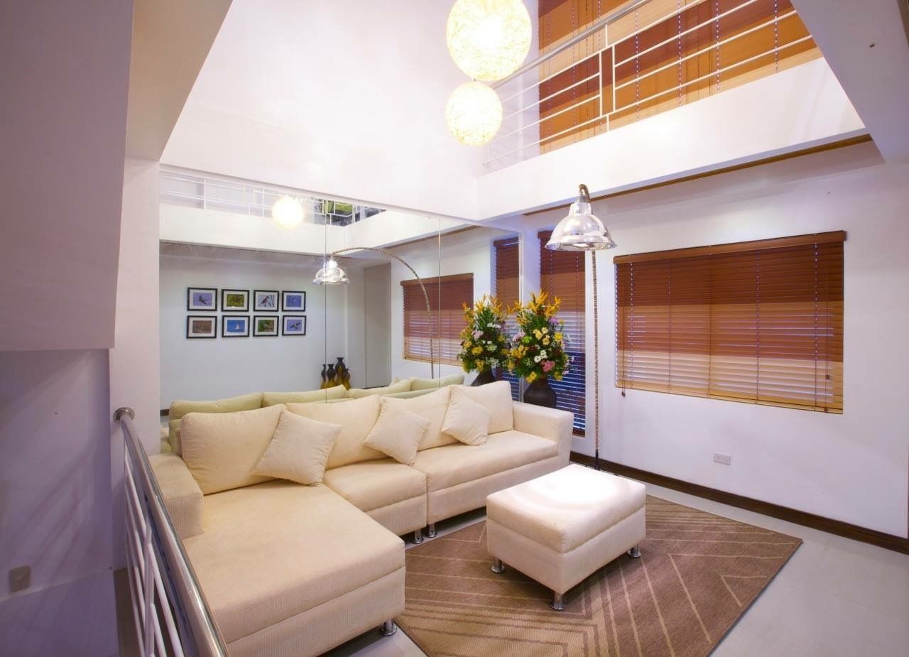 Uma sala de estar moderna com um sofá seccional bege, um banquinho combinando e piso de madeira. Há duas luminárias pendentes, uma janela grande com persianas e uma parede com quadros.