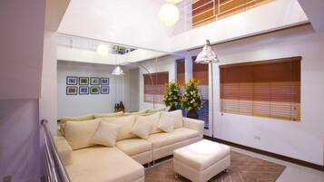 Uma sala de estar moderna com um sofá seccional bege, um banquinho combinando e piso de madeira. Há duas luminárias pendentes, uma janela grande com persianas e uma parede com quadros.