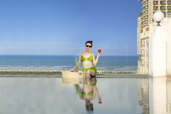 Uma pessoa de biquíni verde sentada à beira da piscina, com uma cesta de vime e uma bebida vermelha, com vista para uma praia e o oceano.