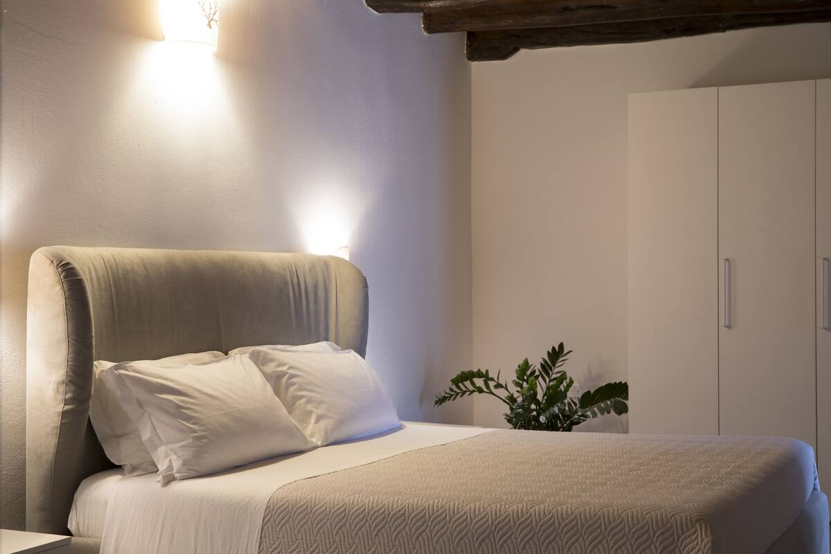 Quarto com cama, mesa de cabeceira, planta e guarda-roupa.