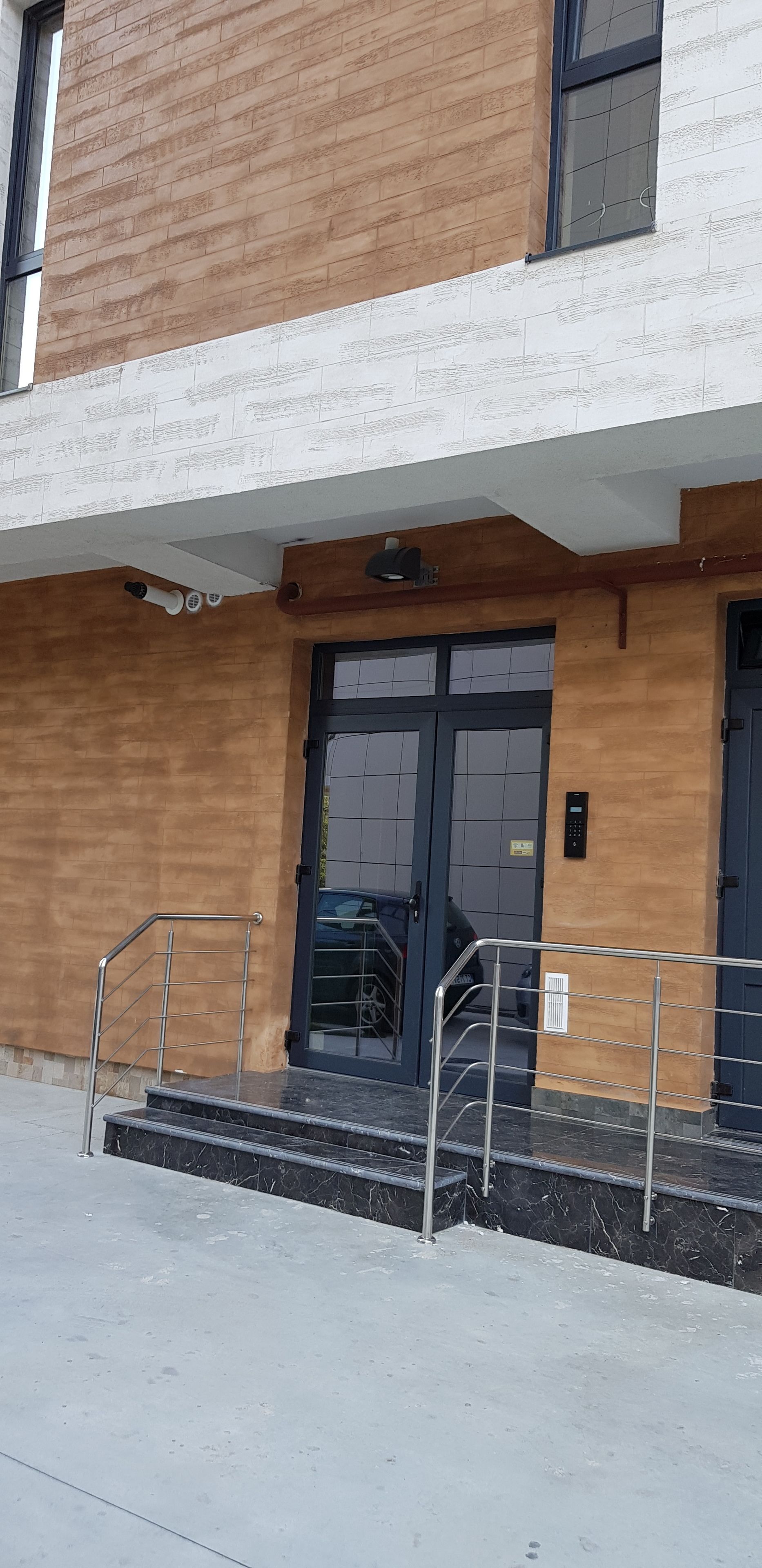 Entrada de um edifício com porta preta, corrimão de metal e um degrau que leva à entrada.