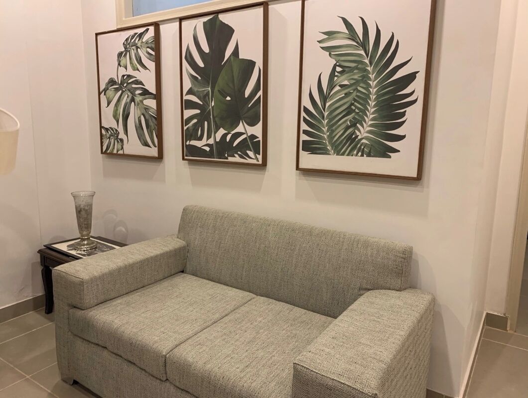 Uma sala de estar com um sofá cinzento, uma mesa pequena com um vaso e três gravuras botânicas emolduradas na parede.
