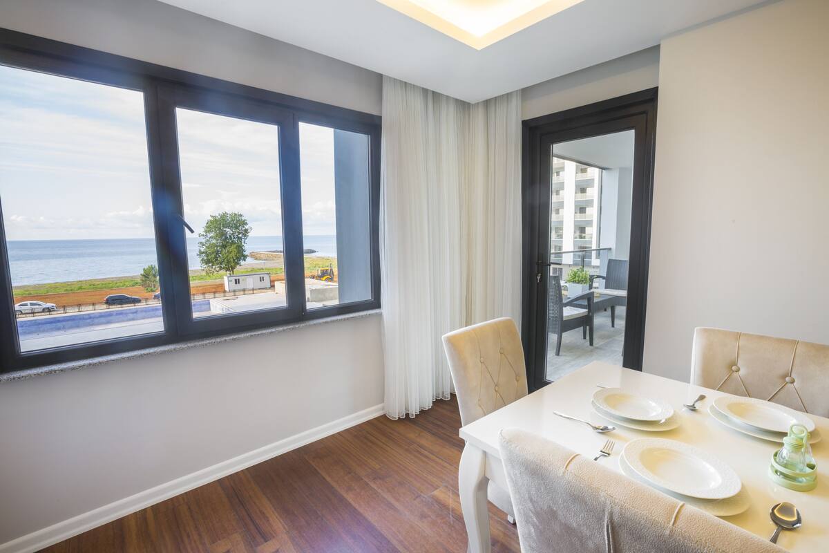 Uma área de refeições com uma mesa posta para quatro pessoas, vista para o mar e uma varanda com uma cadeira.
