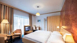Quarto de hotel com uma cama grande, uma escrivaninha com televisão, uma cadeira e um abajur.