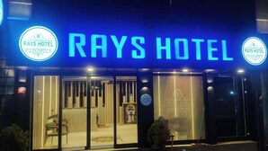 Exterior do HOTEL RAYS com sinalização iluminada, entrada de vidro e interior visível através das portas.