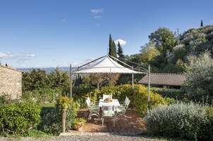 Um alpendre coberto com mesa e cadeiras, rodeado por vegetação exuberante e um edifício de pedra.