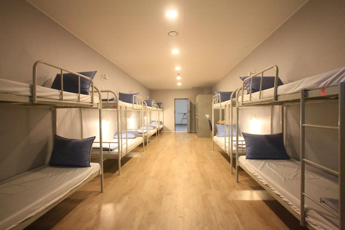 Quarto com beliches dispostos em duas fileiras, piso de madeira e uma porta no final do aposento.