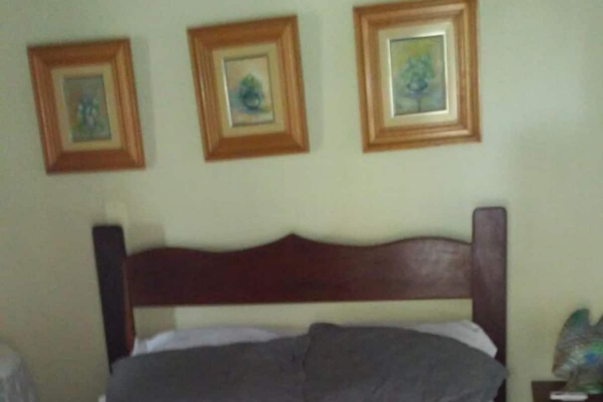 Uma cama com cabeceira de madeira, um edredom cinzento e um cobertor castanho. Duas molduras com quadros estão penduradas na parede acima da cama.
